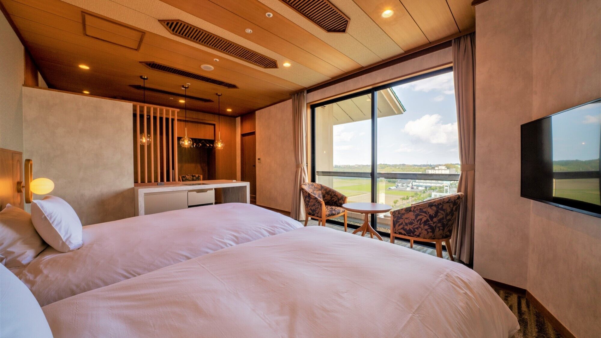 Quarto de hotel com uma cama grande, uma escrivaninha, duas cadeiras, uma televisão e uma janela ampla com vista.
