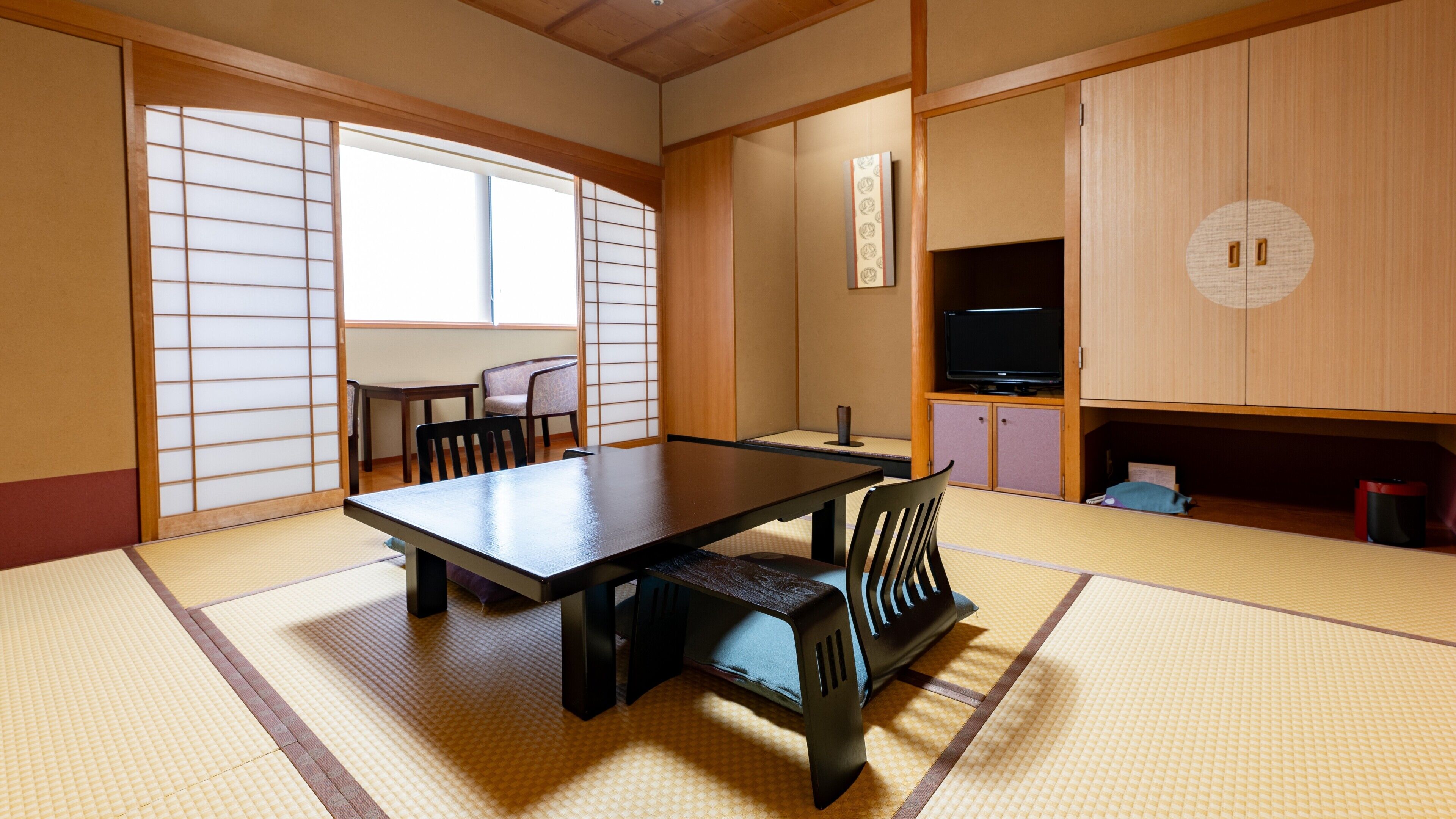 Um quarto japonês tradicional com tatames, uma mesa baixa, cadeiras, uma televisão e portas deslizantes.