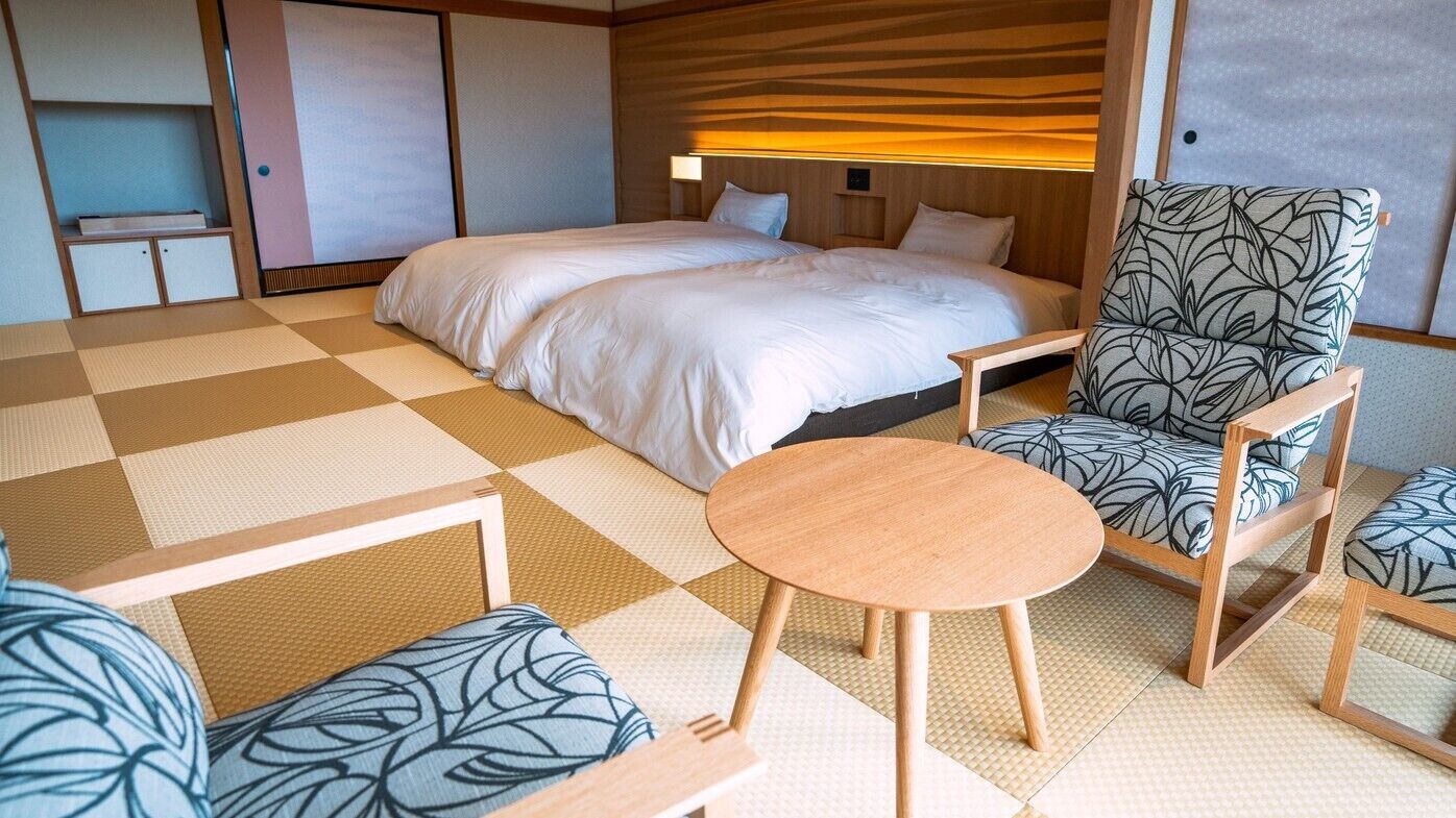 Quarto de hotel com cama, duas cadeiras, uma mesinha e um guarda-roupa.