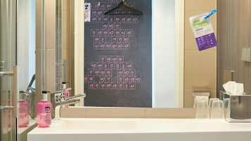 Casa de banho com um espelho grande, uma pia e zona de duche. Há produtos de higiene na bancada da pia e um porta-toalhas acima do espelho.