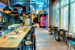 Um restaurante moderno com uma longa mesa comunitária, diversas luminárias pendentes e vista para a cidade através de amplas janelas.