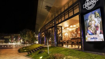 Um edifício moderno com fachada de vidro, área de estar externa e um anúncio proeminente no lado direito.