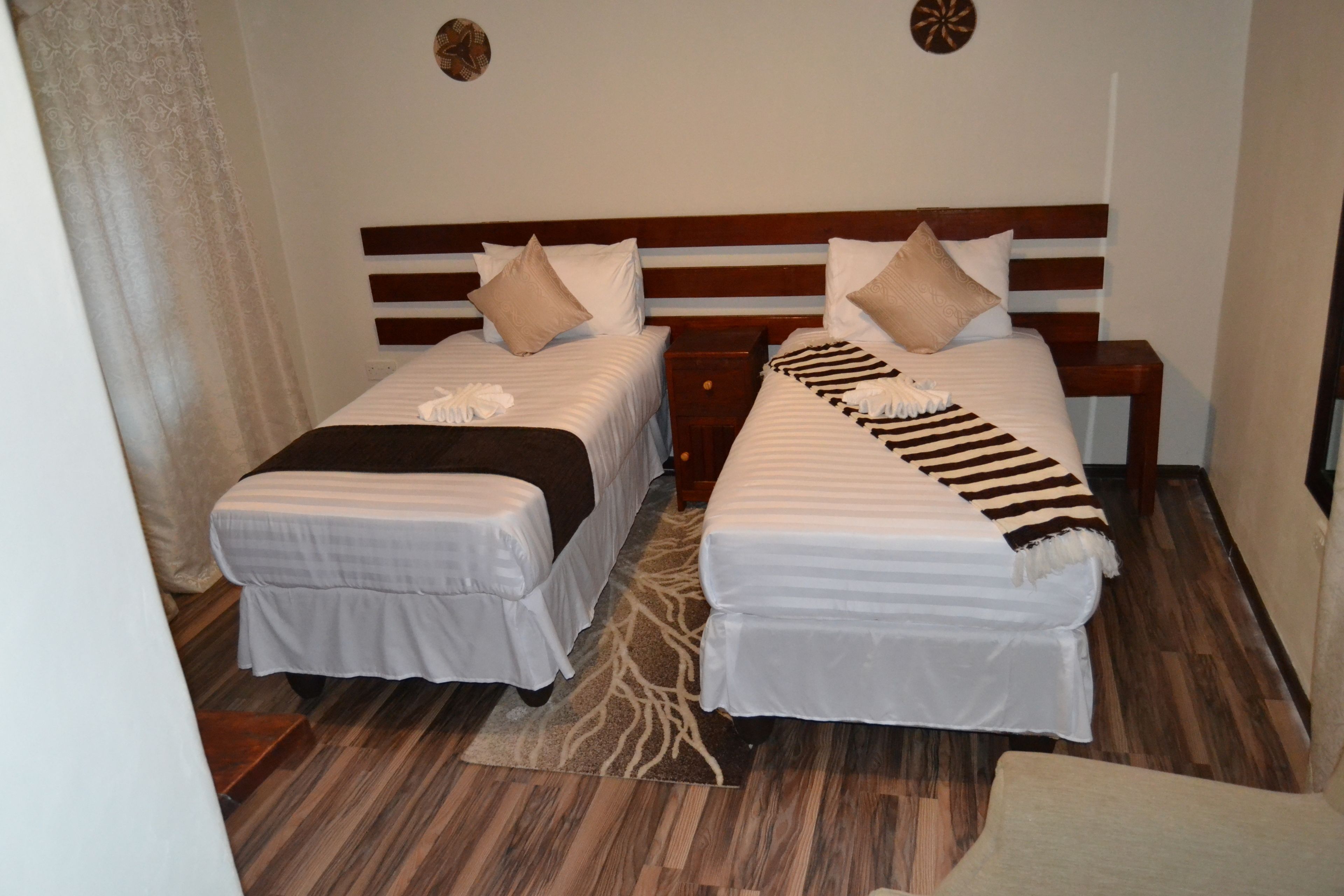 Quarto de hotel com duas camas de solteiro, cabeceira de madeira, pequena mesa de cabeceira e um objeto decorativo na parede.