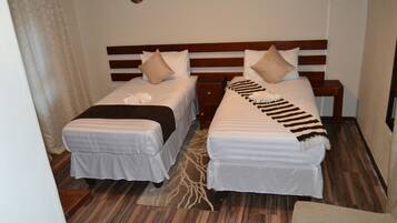 Quarto de hotel com duas camas de solteiro, cabeceira de madeira, pequena mesa de cabeceira e um objeto decorativo na parede.