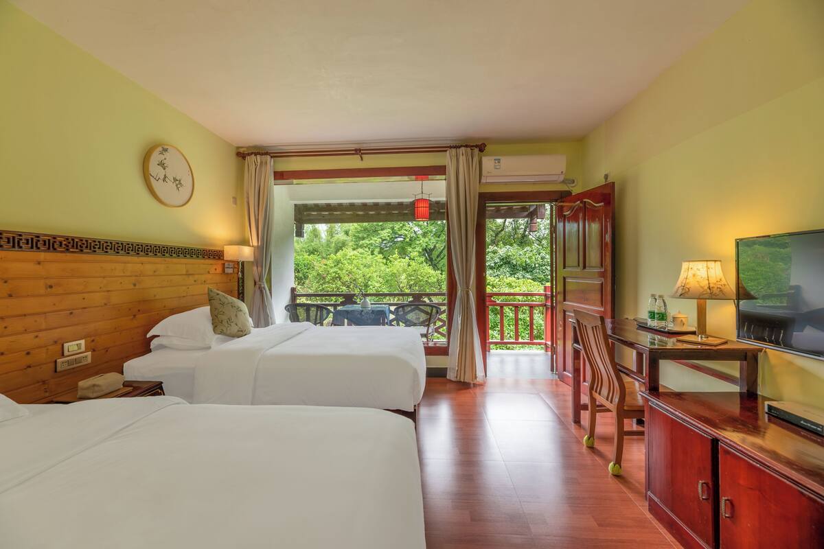 Quarto de hotel com duas camas, televisão, escrivaninha e varanda com vista para o verde.