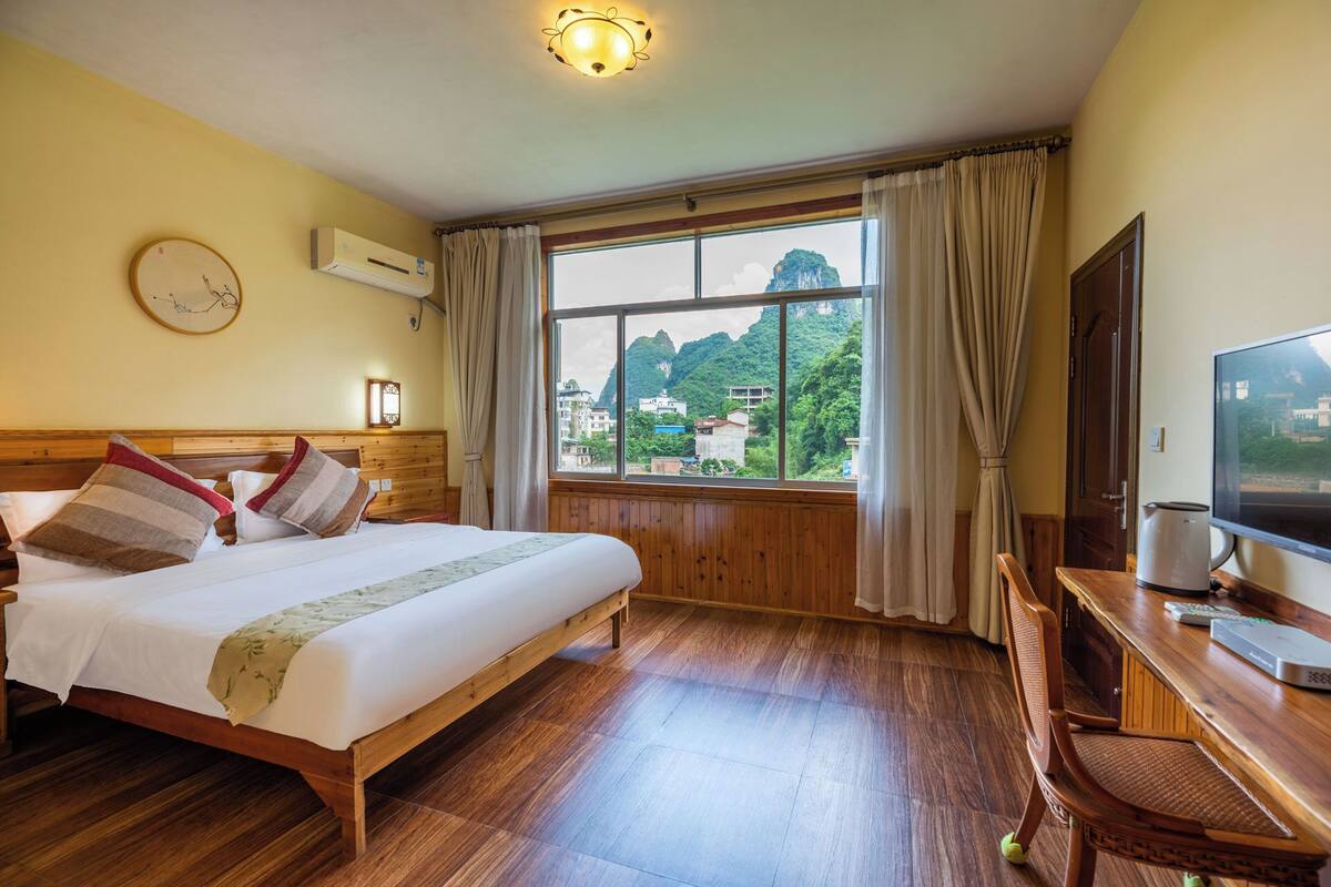 Quarto de hotel com cama, televisão, escrivaninha e janela com vista para uma paisagem montanhosa.