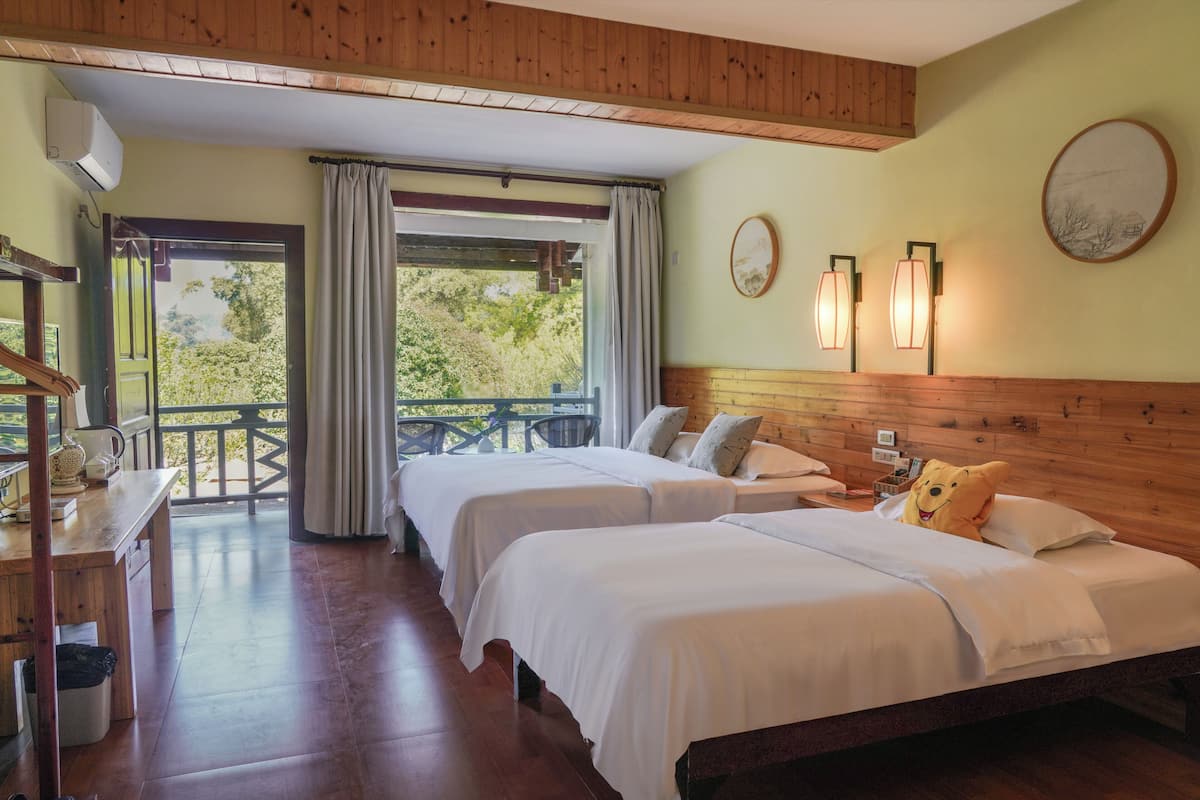Quarto com duas camas, teto de madeira, janela com cortinas, mesa com chaleira e luminárias fixadas na parede.