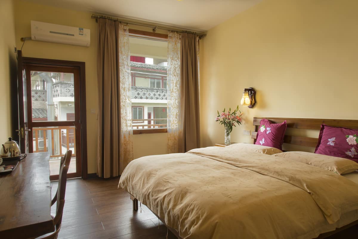 Quarto com cama grande, mesa de cabeceira de madeira, escrivaninha com cadeira e janela com cortinas.