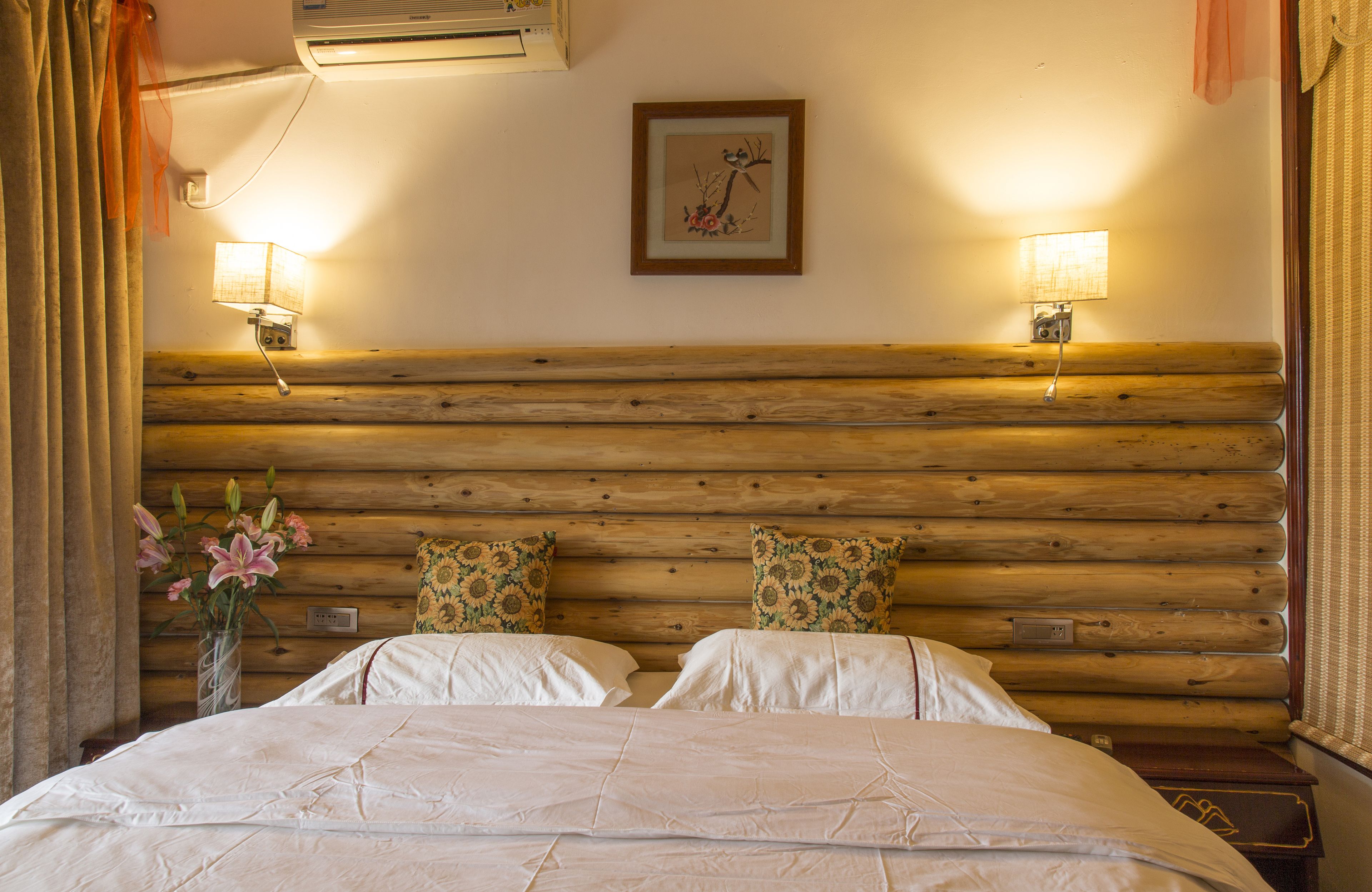 Cama com cabeceira de madeira, dois travesseiros, um quadro emoldurado e duas luminárias fixadas na parede.