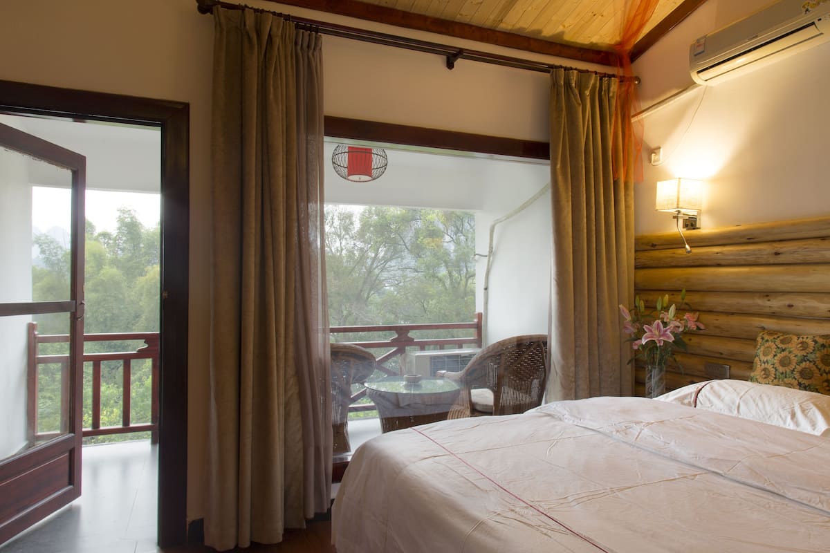 Quarto com cama, janela com cortinas, mesa e cadeiras, e vista para o exterior.