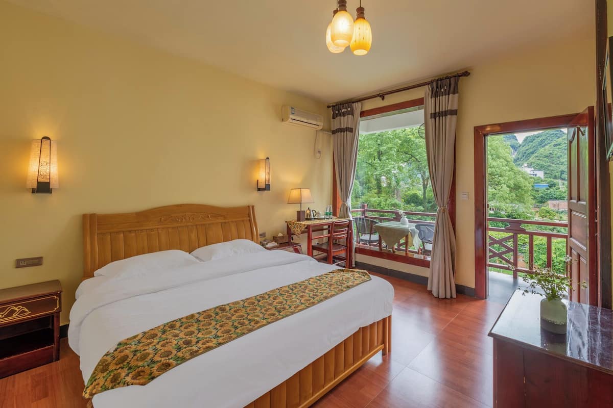 Quarto de hotel com cama, mesa de cabeceira, luminária e varanda com mesa e cadeiras.