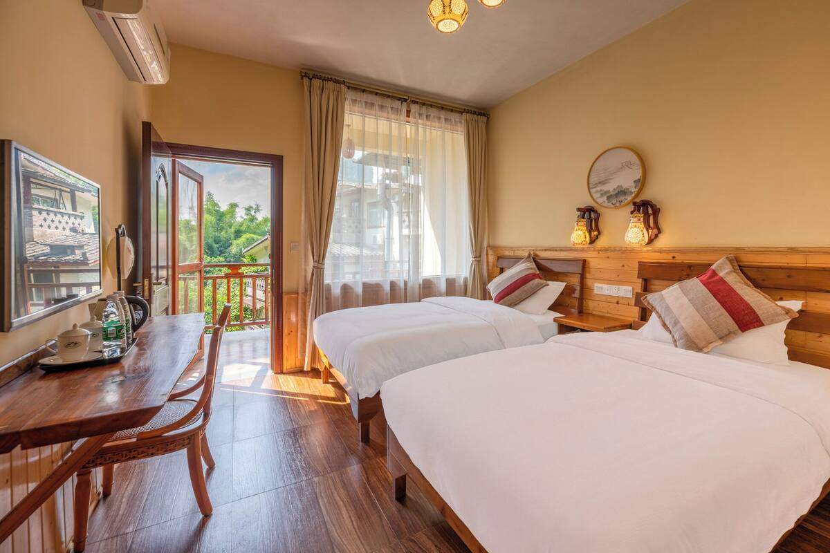 Quarto de hotel com duas camas, uma escrivaninha de madeira com cadeira, uma mesa com uma bandeja de objetos e uma varanda com vista para o verde.