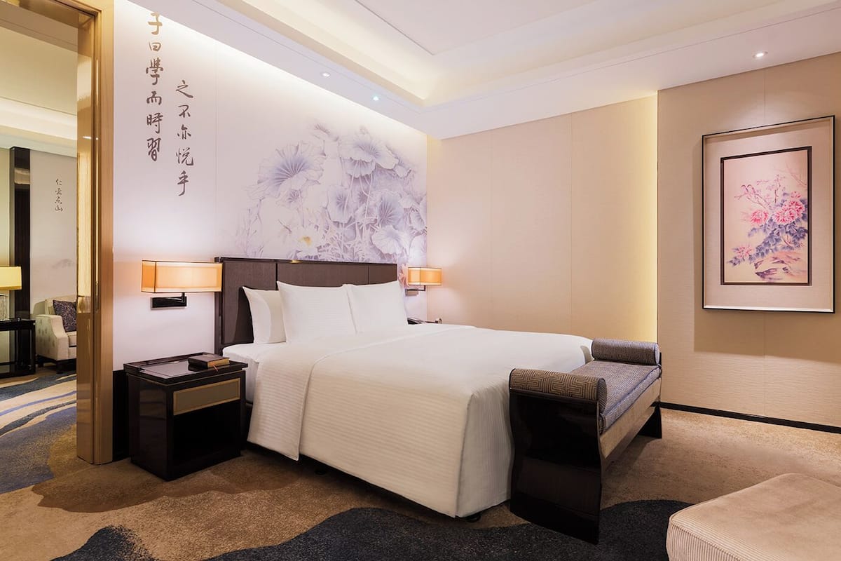 Quarto de hotel com uma cama grande, duas mesas de cabeceira, um criado-mudo, um sofá e uma parede com um mural floral.