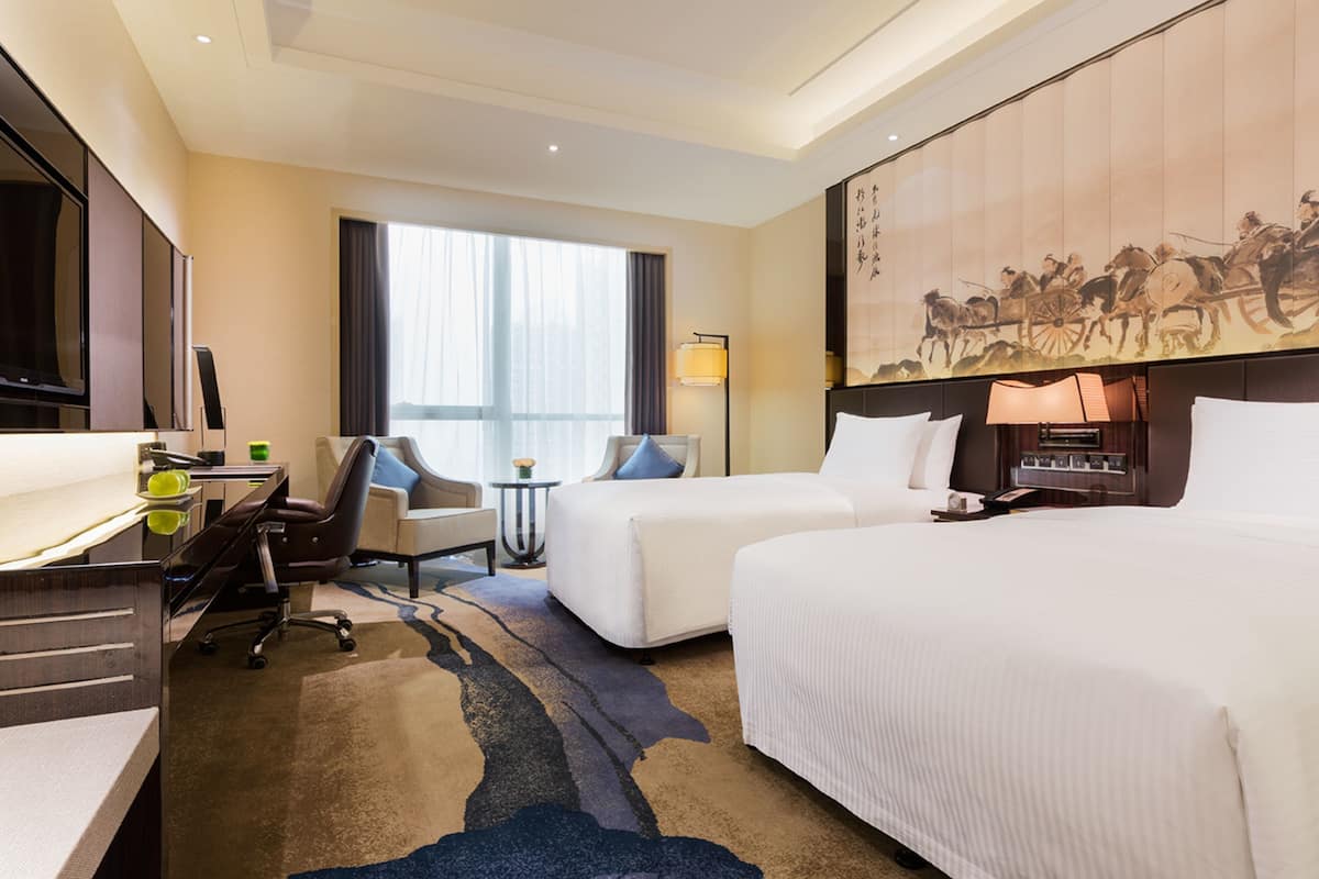 Quarto de hotel com duas camas, uma mesa, uma cadeira, uma televisão e uma janela grande.