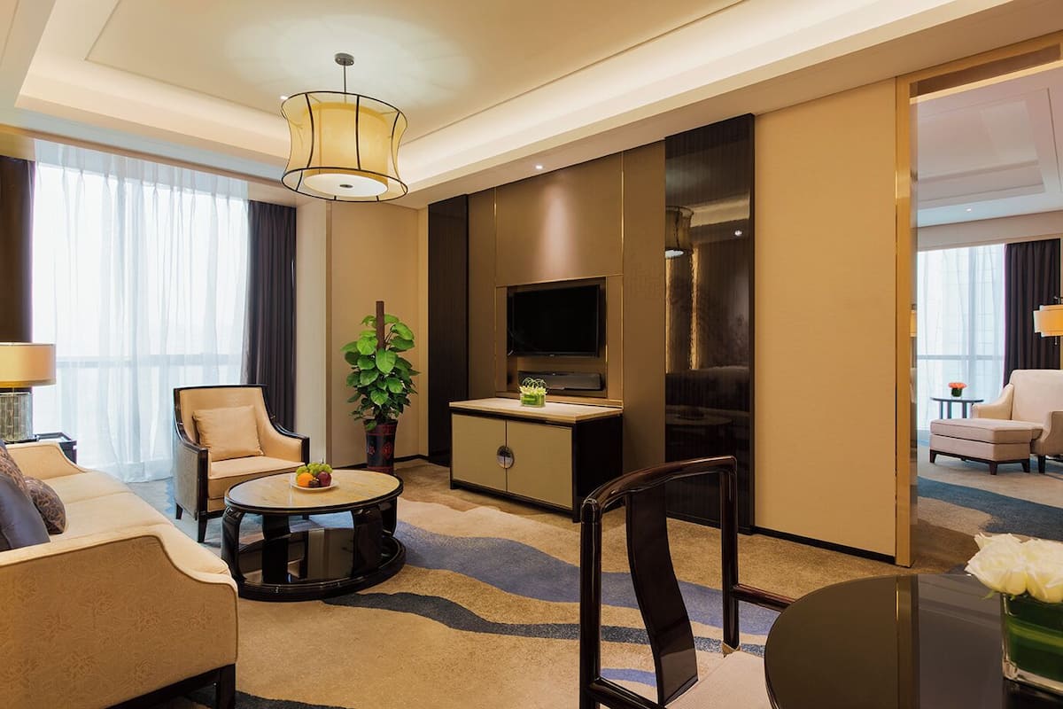 Quarto de hotel moderno com sofá, poltrona, mesa de centro redonda, televisão e planta em vaso.