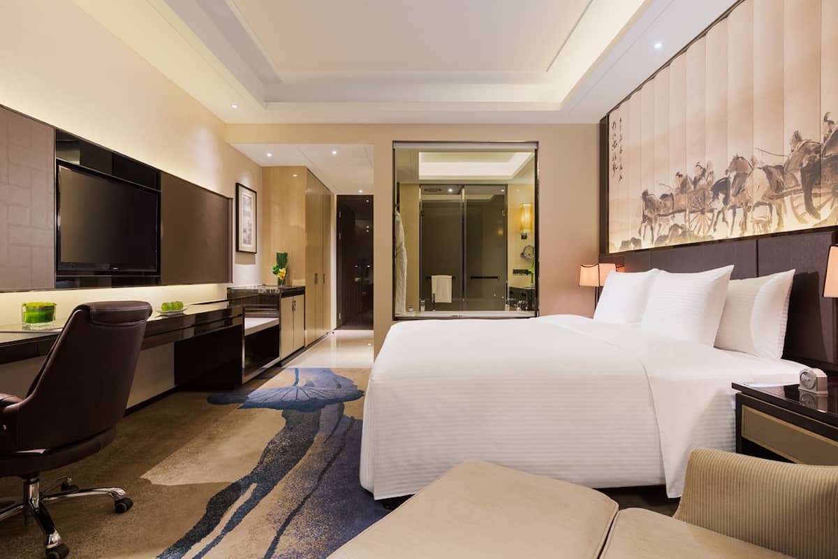 Quarto de hotel com uma cama grande, uma mesa com televisão de tela plana, uma cadeira e um banheiro visível por uma porta aberta.