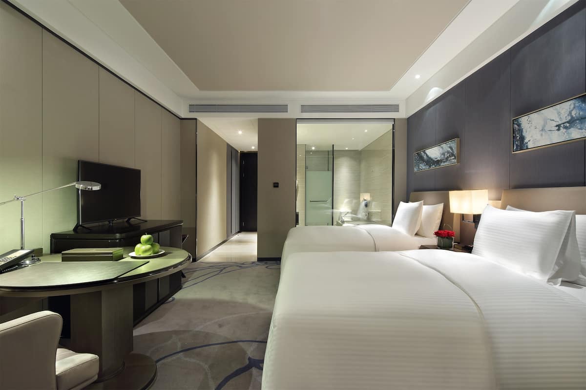 Quarto de hotel moderno com uma cama grande, uma escrivaninha com cadeira, televisão e banheiro visível através de uma porta de vidro.
