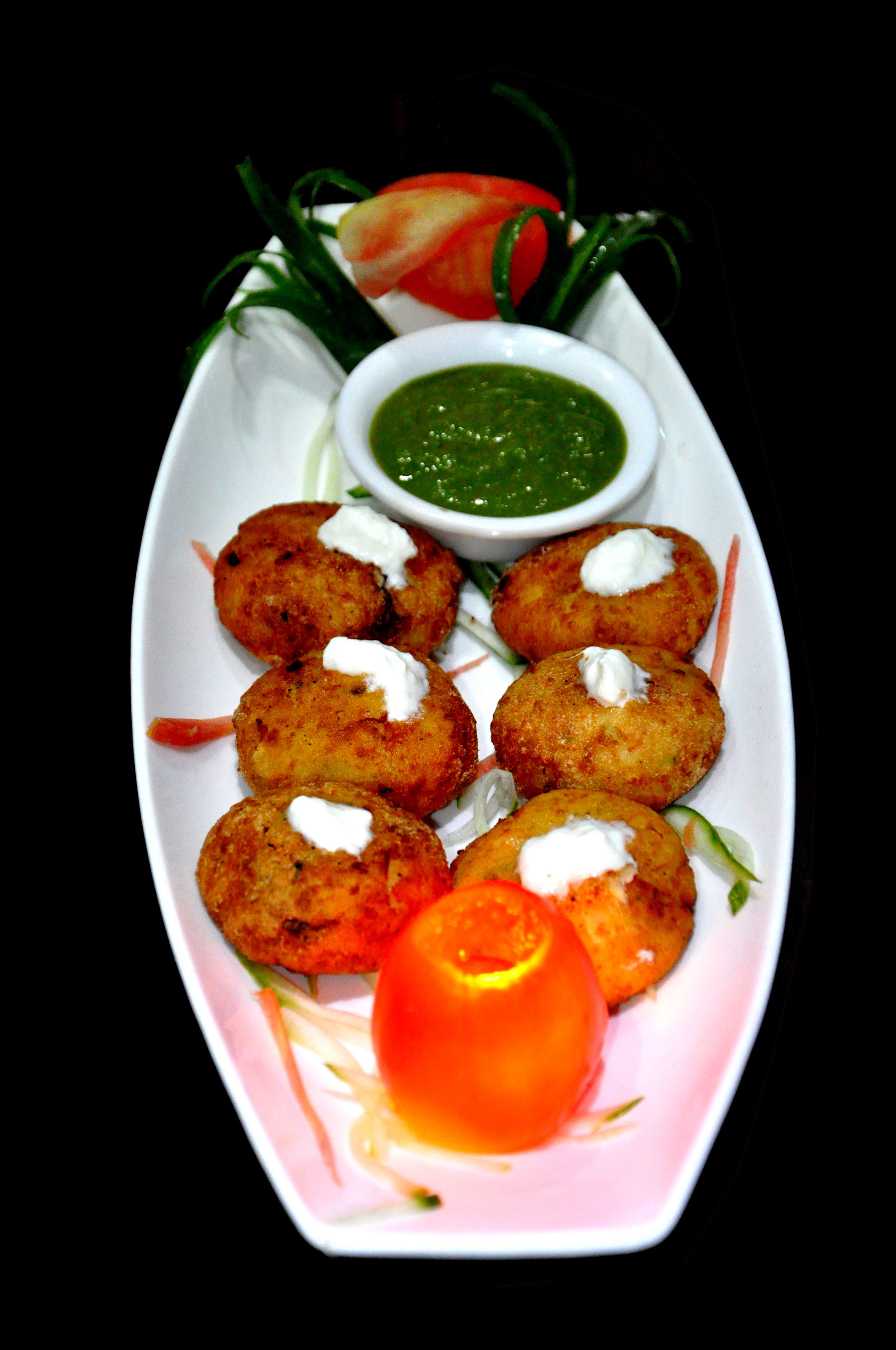 Um prato de comida com seis itens redondos cobertos por um molho branco, uma fatia de tomate e uma pequena tigela de molho verde.