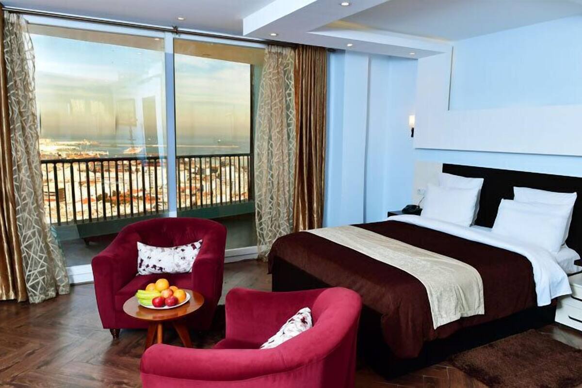 Quarto de hotel com uma cama grande, uma poltrona vermelha, uma mesinha com frutas e uma varanda com vista.