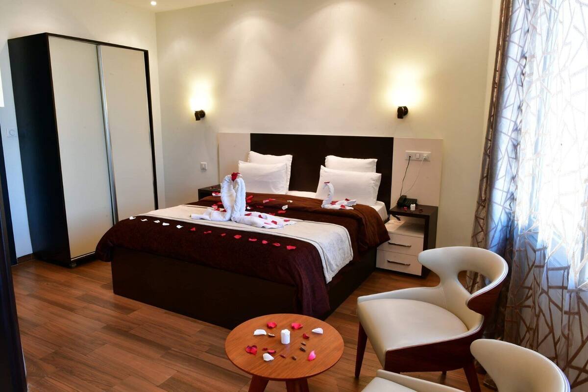 Quarto de hotel com uma cama, duas mesas de cabeceira, uma cadeira, uma pequena mesa com pétalas de rosa e um guarda-roupa.