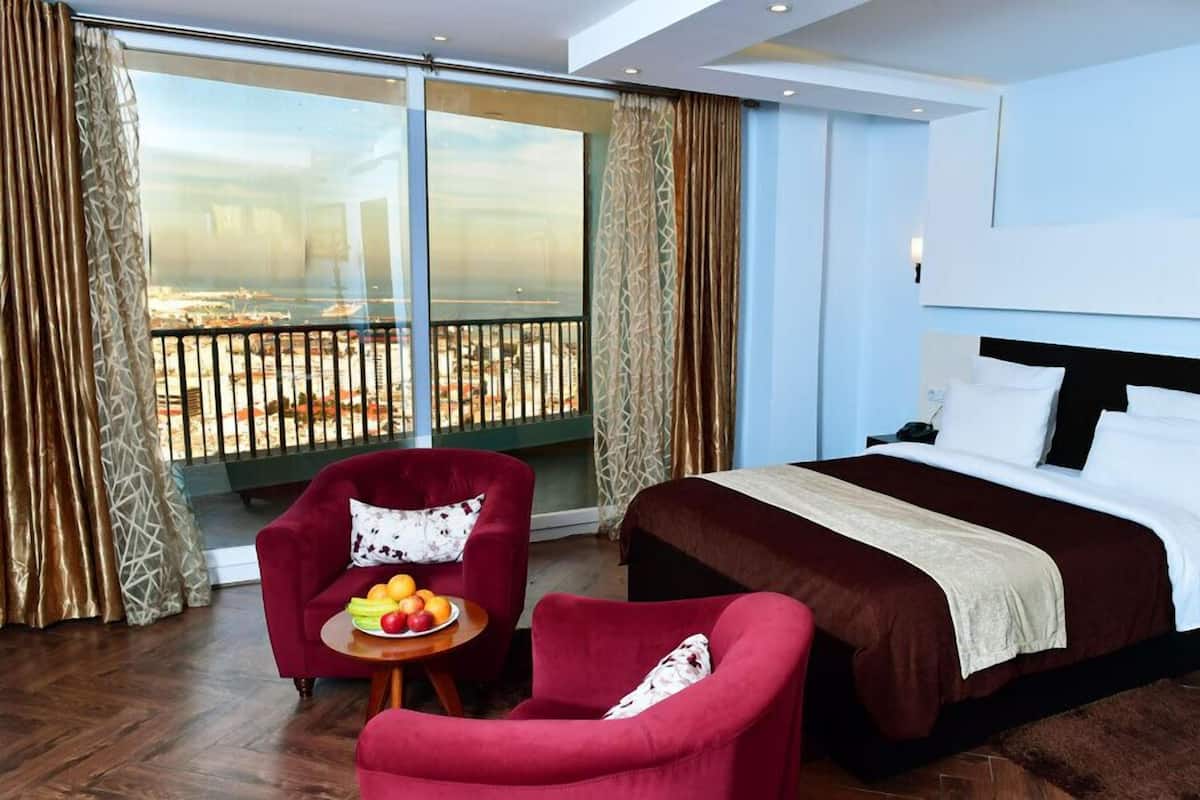 Quarto de hotel com uma cama grande, duas poltronas vermelhas, uma mesinha com frutas e varanda com vista.