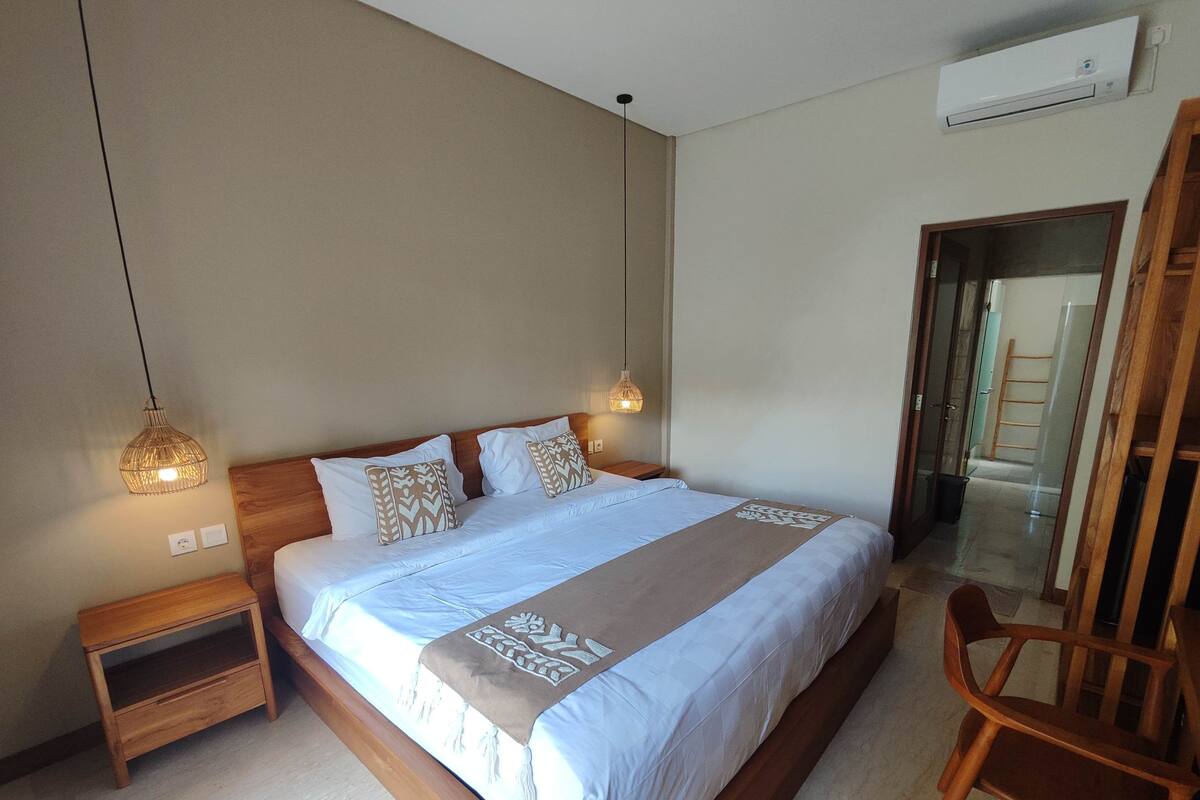 Quarto com cama de madeira, mesas de cabeceira, uma cadeira e luminária pendente.