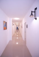 Um corredor longo e bem iluminado, com paredes brancas e chão de azulejos, que termina numa porta azul.