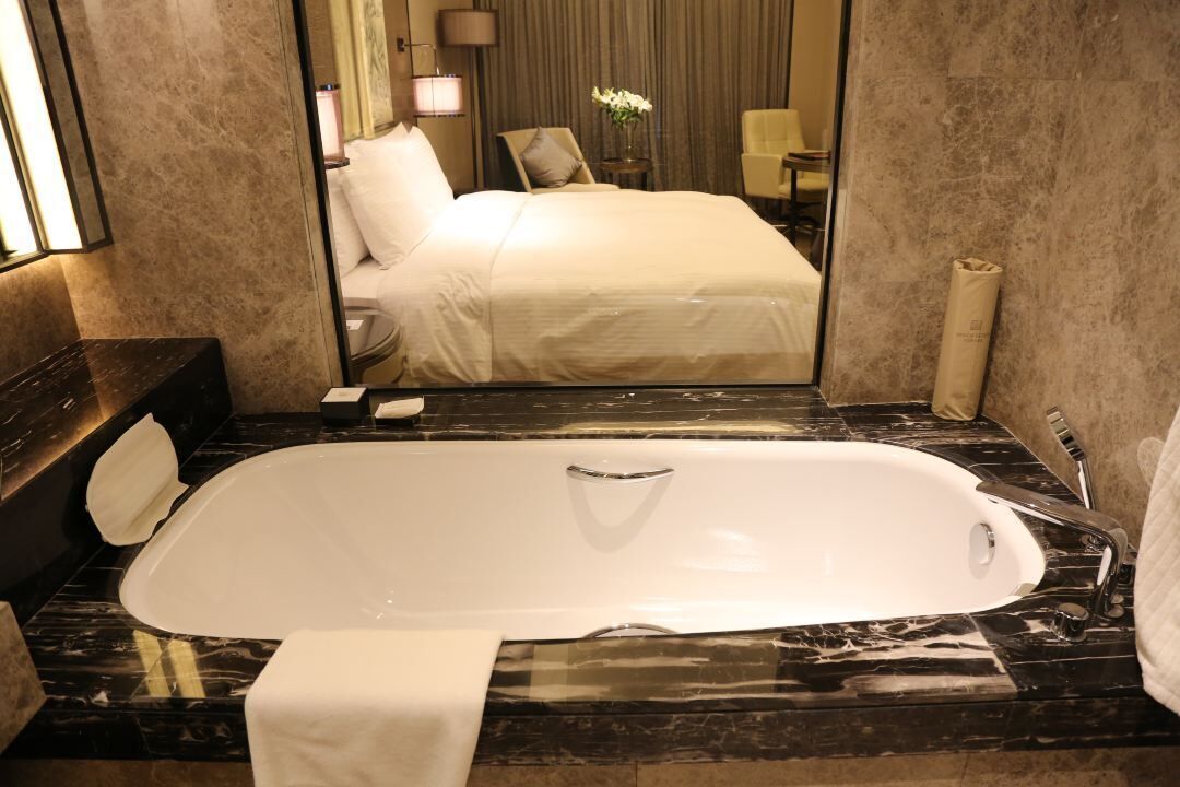 Casa de banho moderna com uma banheira grande, bancadas de mármore e um espelho refletindo um quarto com cama e cortinas.