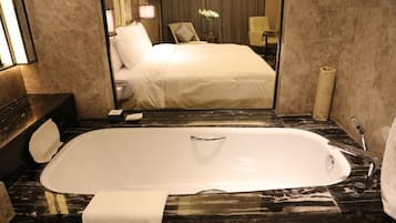 Casa de banho moderna com uma banheira grande, bancadas de mármore e um espelho refletindo um quarto com cama e cortinas.