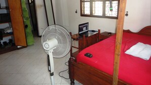 Quarto com cama, escrivaninha, laptop, ventoinha, espelho e janela com grades.