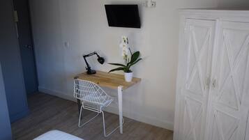 Um quarto pequeno com uma escrivaninha, uma cadeira, uma orquídea em vaso, uma TV fixada na parede e um armário branco.