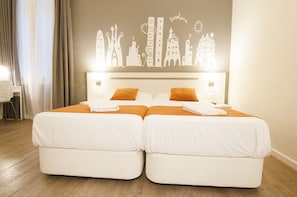 Quarto de hotel com duas camas, roupa de cama branca, almofadas decorativas laranja, uma pintura de paisagem urbana e piso de madeira.