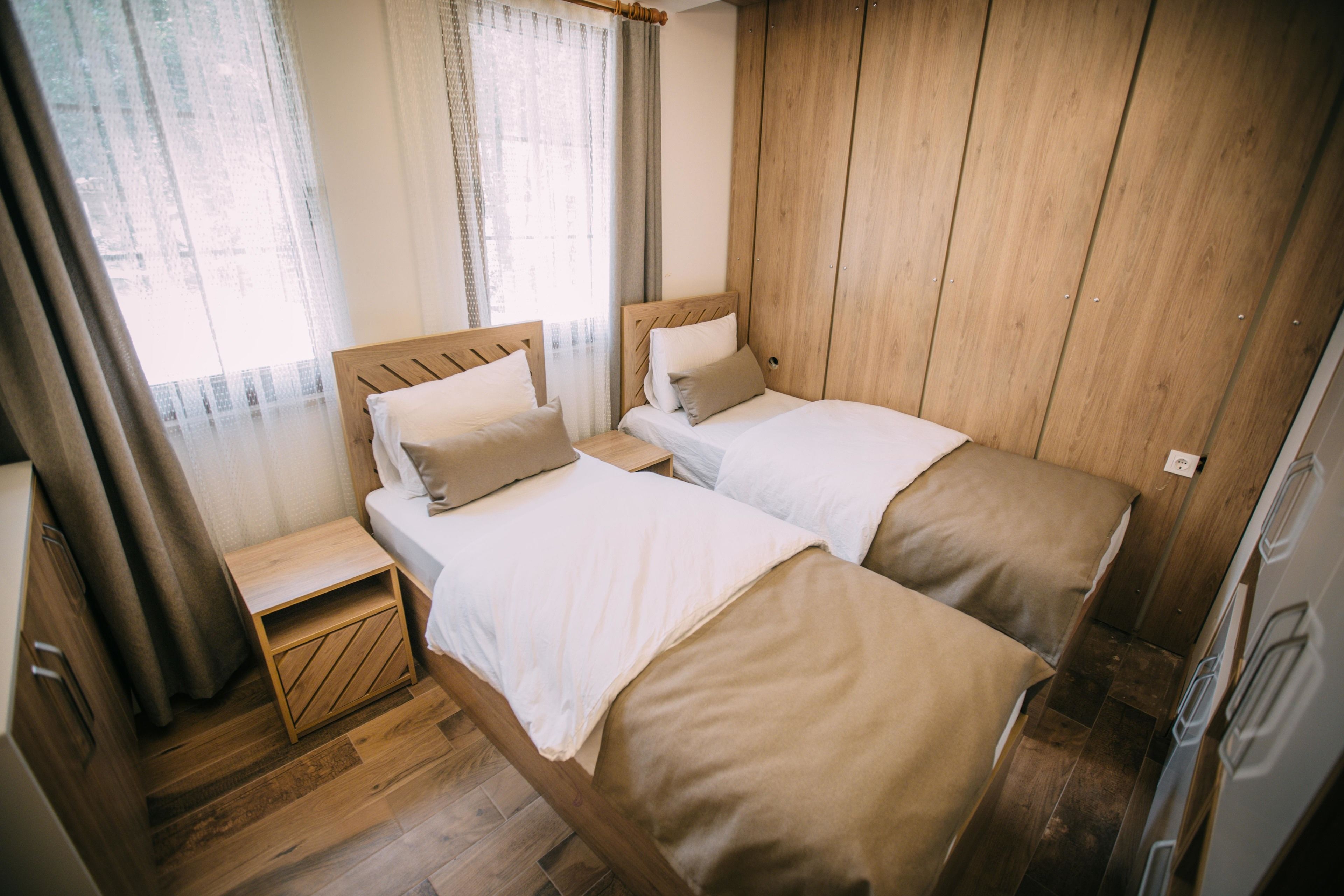 Quarto compacto com duas camas, paredes de madeira, uma pequena mesa de cabeceira e uma janela com cortinas.