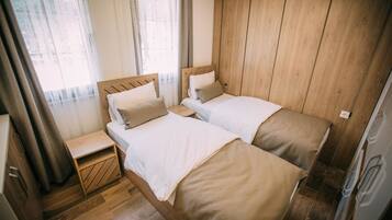 Quarto compacto com duas camas, paredes de madeira, uma pequena mesa de cabeceira e uma janela com cortinas.