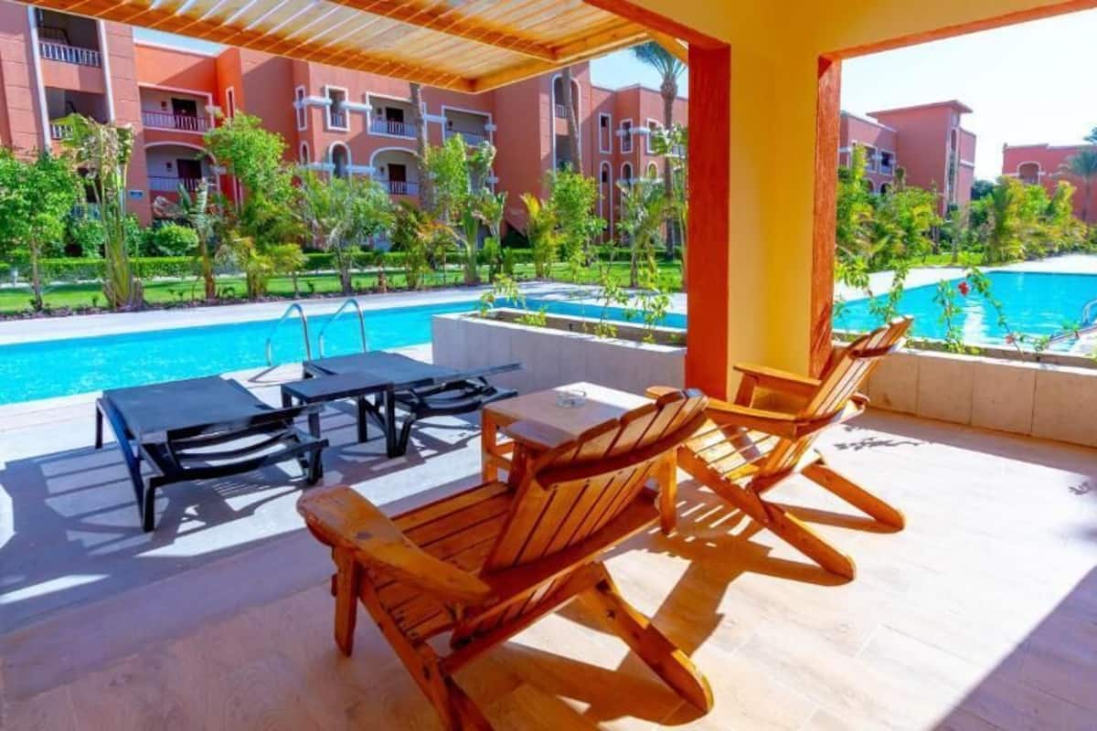 Uma área à beira da piscina com espreguiçadeiras de madeira e uma mesa, com vista para uma piscina e um edifício com varandas.