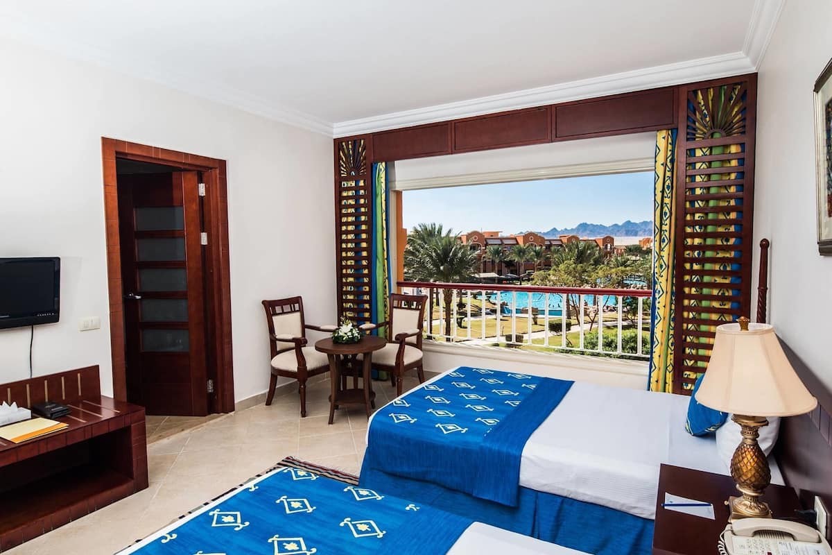 Quarto de hotel com varanda, uma cama com colcha azul, uma escrivaninha com cadeira e uma televisão.