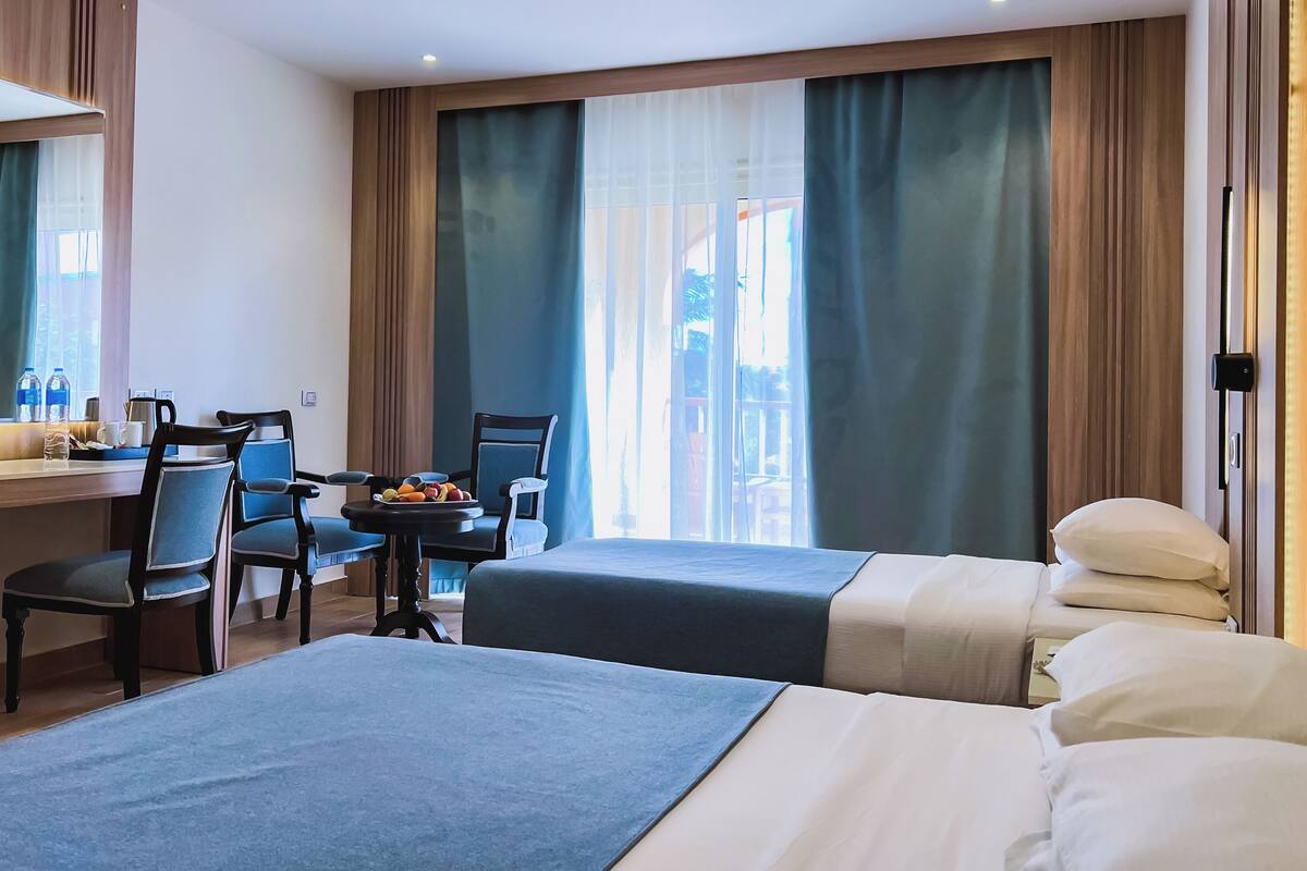 Quarto de hotel com duas camas, uma escrivaninha com cadeiras, uma mesinha com fruta e grandes janelas com cortinas.