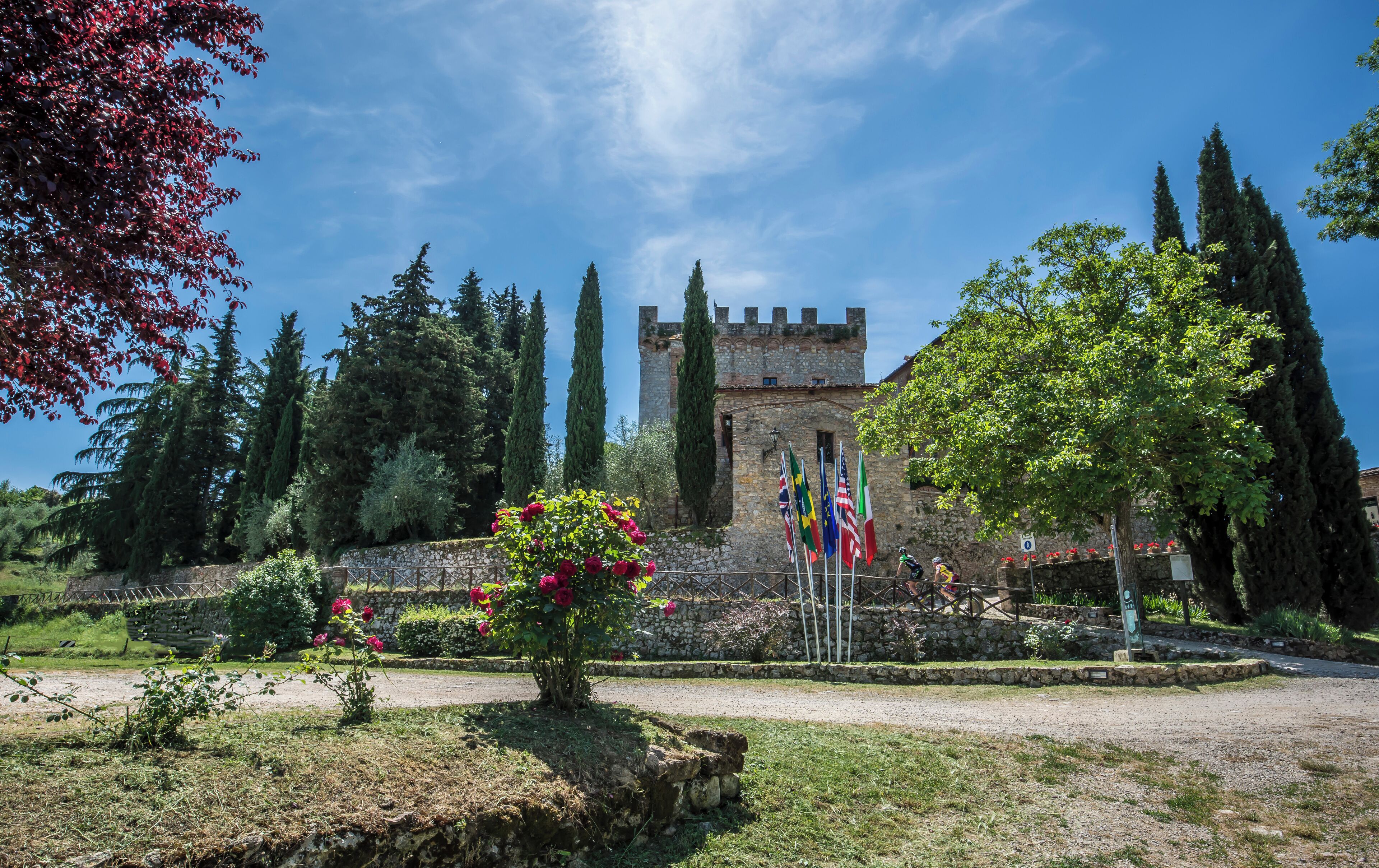 Um castelo histórico com uma parede de pedra, exibição de bandeiras e um jardim bem cuidado, com árvores e flores.