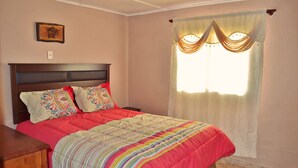 Quarto com cama, mesa de cabeceira, cabeceira de madeira, janela com cortinas e um quadro na parede.