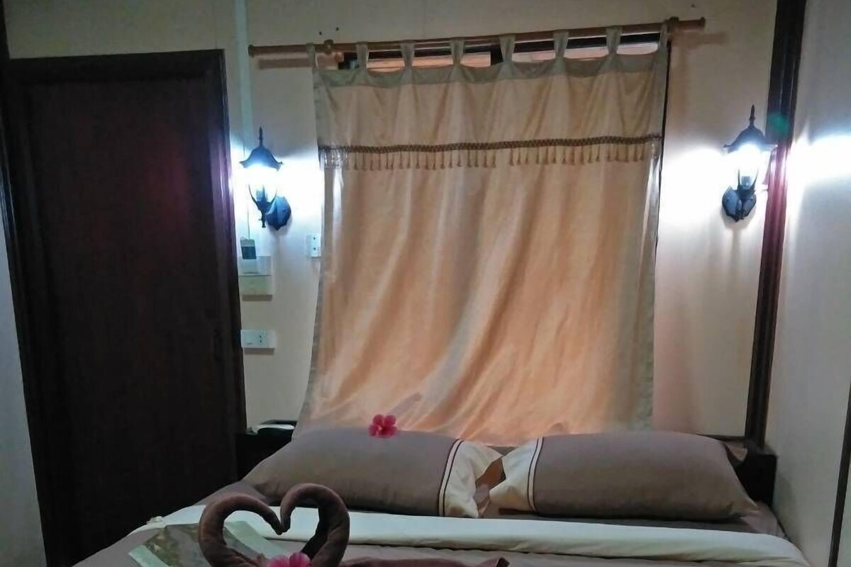 Cama bem arrumada com toalha em formato de coração e flores cor-de-rosa, cortina bege e duas luminárias fixadas na parede.