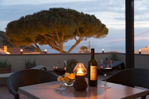 Uma mesa com duas taças de vinho, uma vela e uma garrafa de vinho num terraço ao ar livre com vista para uma árvore e o mar.