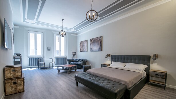 Um quarto espaçoso com uma cama grande, um sofá de couro e uma mesa de jantar. Duas janelas oferecem luz natural, e o quarto apresenta um design moderno no teto.
