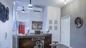 Balcão de recepção com a inscrição "STEAM HOUSE", um laptop, um globo e quadros na parede. Há dois banquetes e um armário vermelho ao fundo.