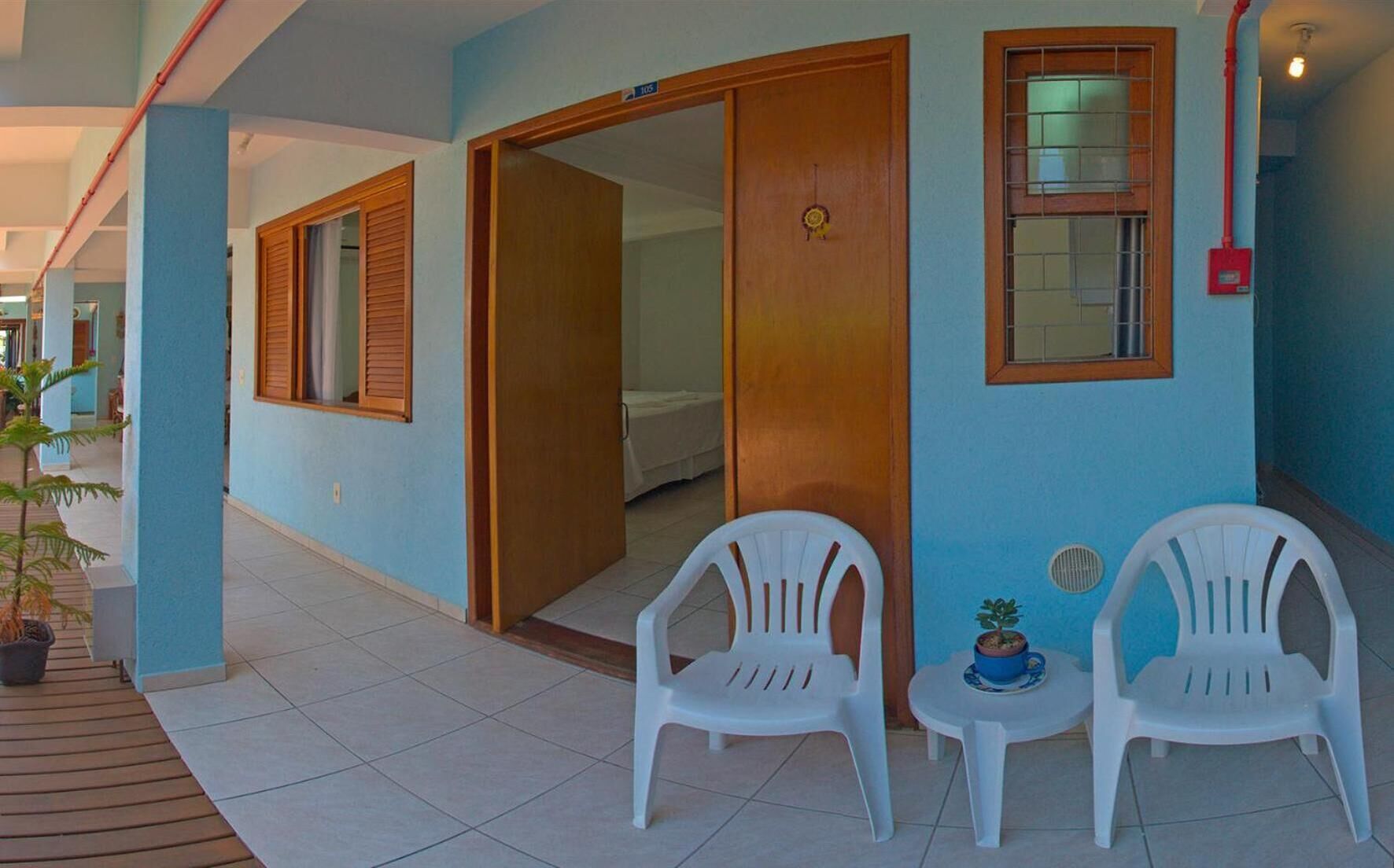 Quarto de hotel com porta deslizante, duas cadeiras de plástico branco, uma mesinha com planta em vaso e janela com grade de metal.