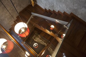 Escadaria moderna com degraus de madeira e guarda-corpo de vidro, que desce para um nível inferior com um candeeiro circular e uma pequena planta.
