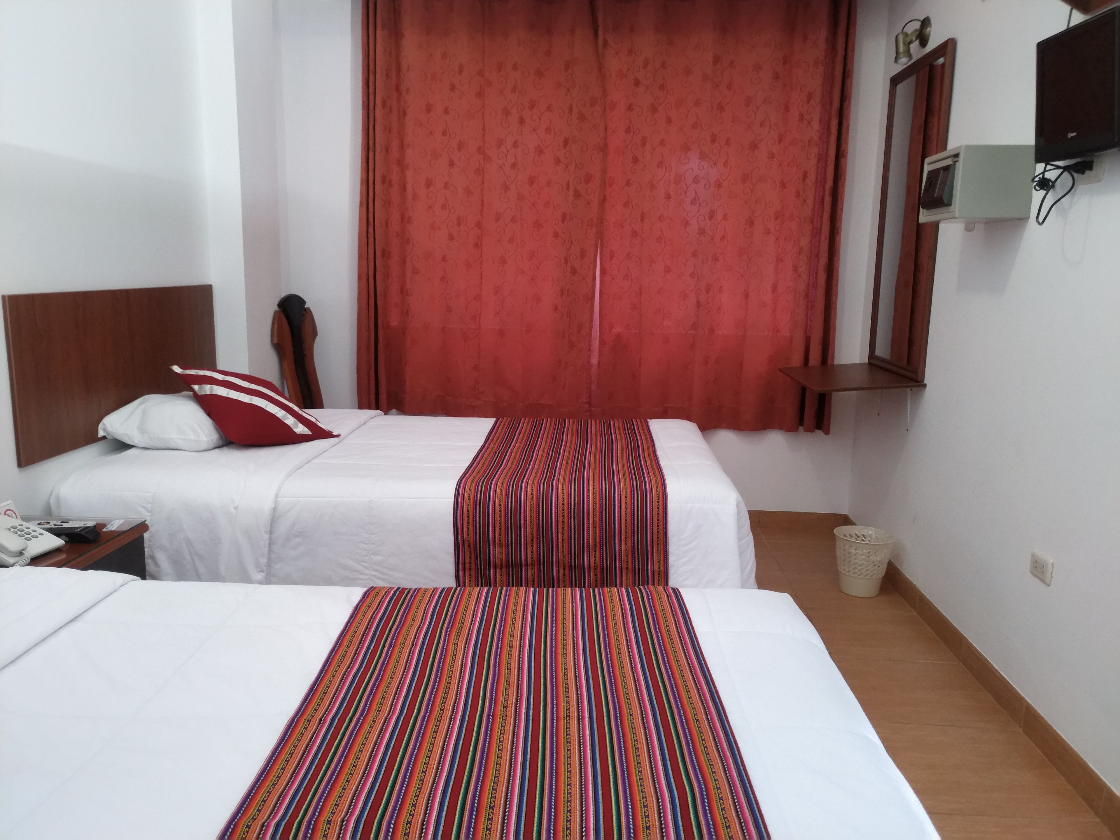 Quarto de hotel com duas camas, cada uma com colcha branca e manta listrada colorida. Possui cabeceira de madeira, um telefone e uma pequena mesa com abajur. Cortina vermelha e um espelho na parede.