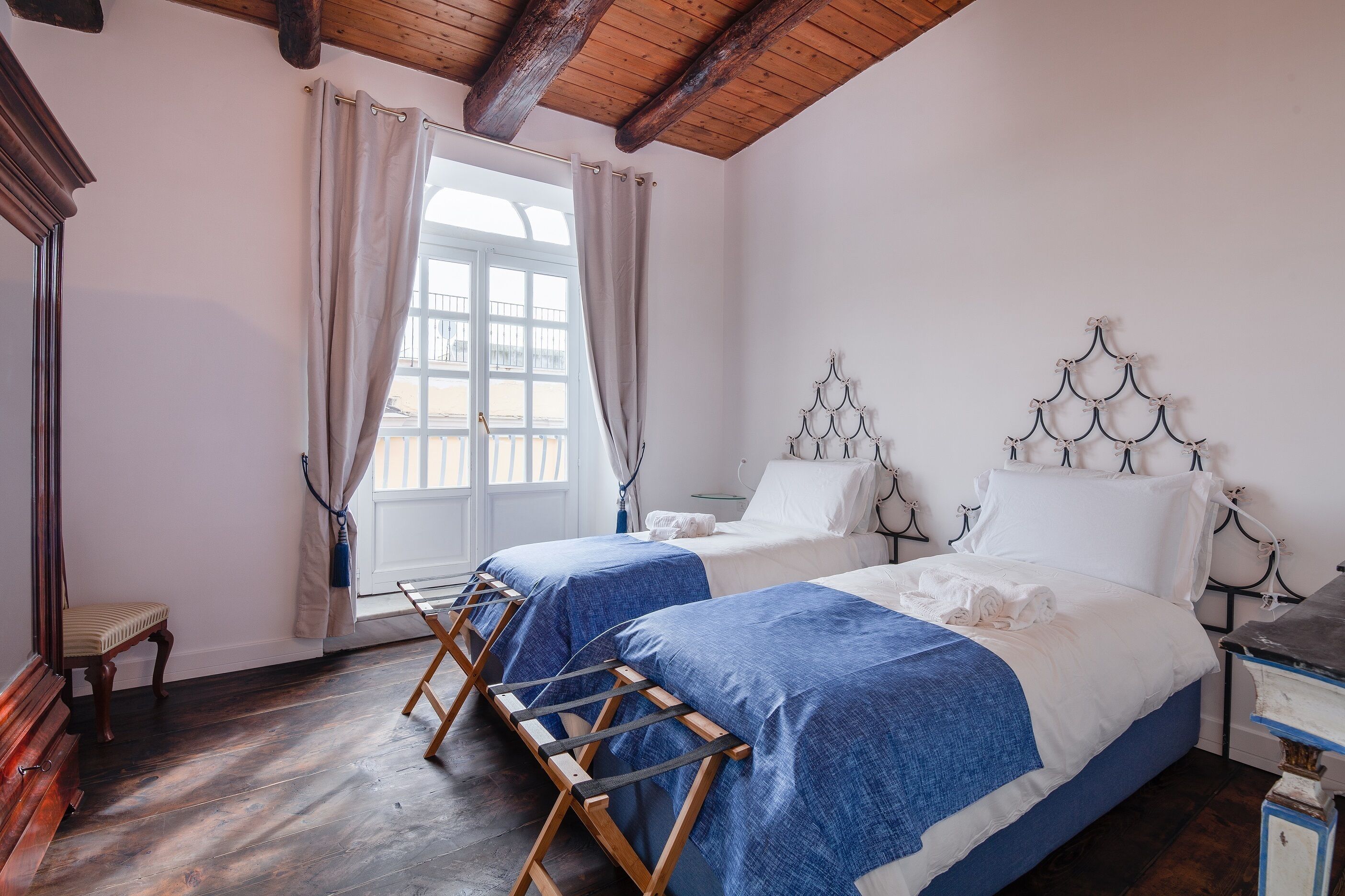 Quarto com duas camas, soalho em madeira, janela com cortinas e teto de madeira.