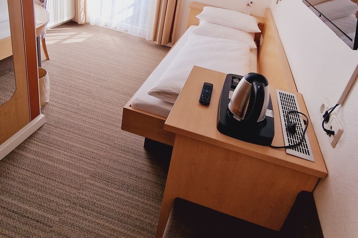 Quarto de hotel com uma cama, uma mesa com um laptop, um comando remoto e uma chaleira.