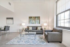 Uma sala de estar moderna com um sofá cinzento, duas poltronas, uma mesa de centro e uma área de jantar com uma mesa de vidro e cadeiras.