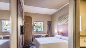 Quarto de hotel com duas camas, uma escrivaninha, uma cadeira, uma televisão, uma janela com vista para edifícios e um banheiro com espelho e pia.